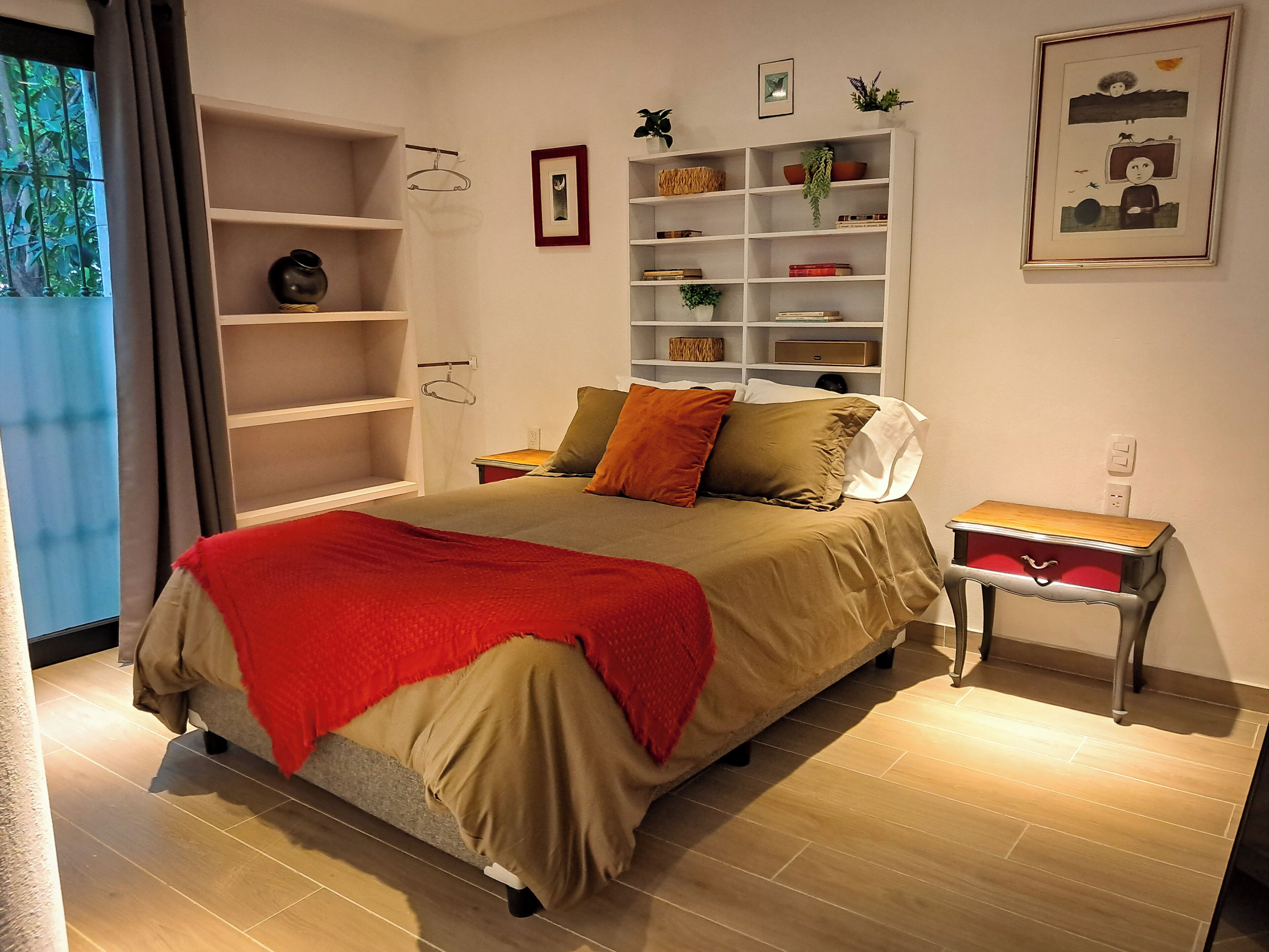 Um quarto com cama, mesa de cabeceira, estante e janela com cortinas.