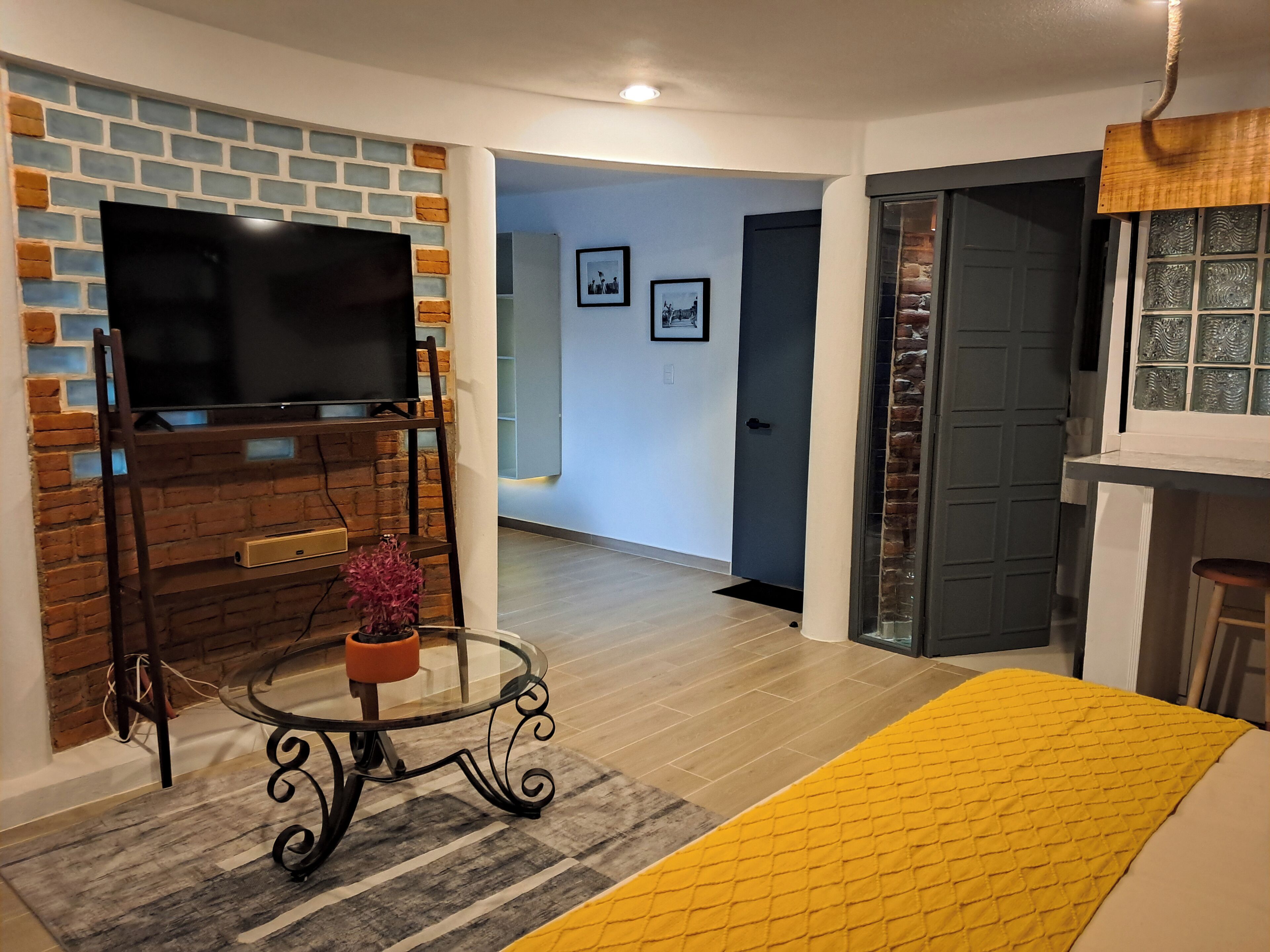 Uma sala de estar com televisão de tela plana fixada em uma parede de tijolos, uma mesa de centro de vidro com uma planta em vaso, e um cobertor amarelo em um sofá.