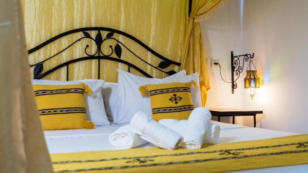 Uma cama com roupa de cama amarela e branca, cabeceira de metal preto e luminária fixada na parede.