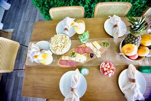 Uma mesa de jantar posta com pratos, guardanapos e diversos alimentos, incluindo queijo, carnes, frutas e petiscos.