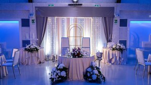 Espaço para recepção de casamento com mesas redondas, cadeiras, arranjos florais e palco com painel de fundo.