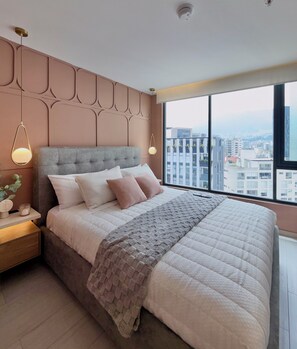Quarto moderno com uma cama grande, mesa de cabeceira, luminária fixada na parede e vista da cidade através de amplas janelas.