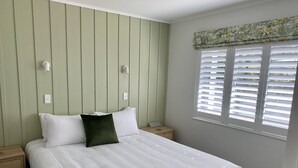 Cama bem arrumada com almofadas brancas e verde-escuro, mesa de cabeceira de madeira com abajur e janela com persianas.