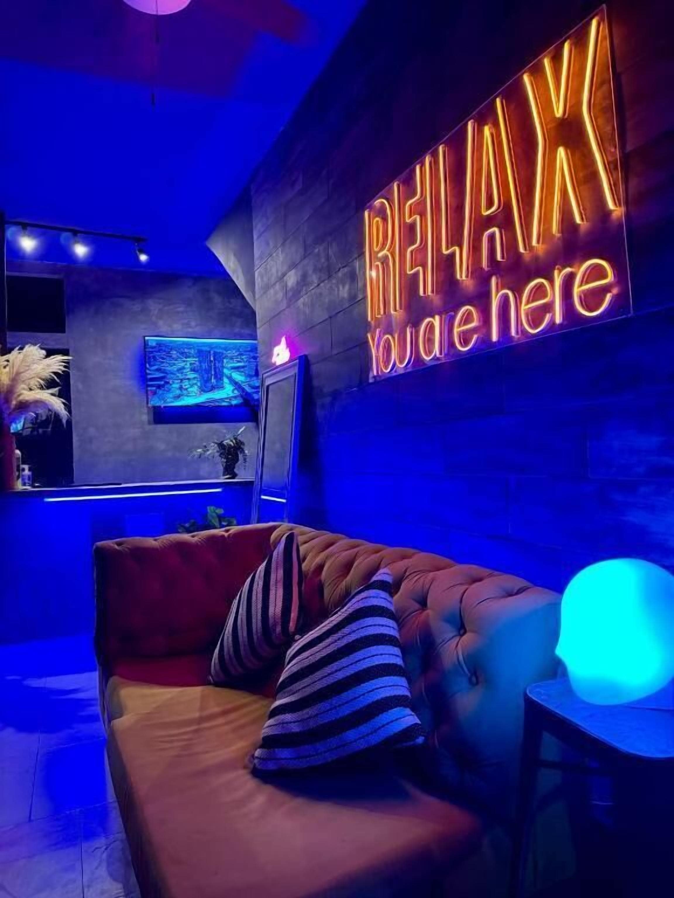Uma área de lounge moderna com um letreiro de neon que diz “RELAX You are here” e um sofá confortável com almofadas listradas.