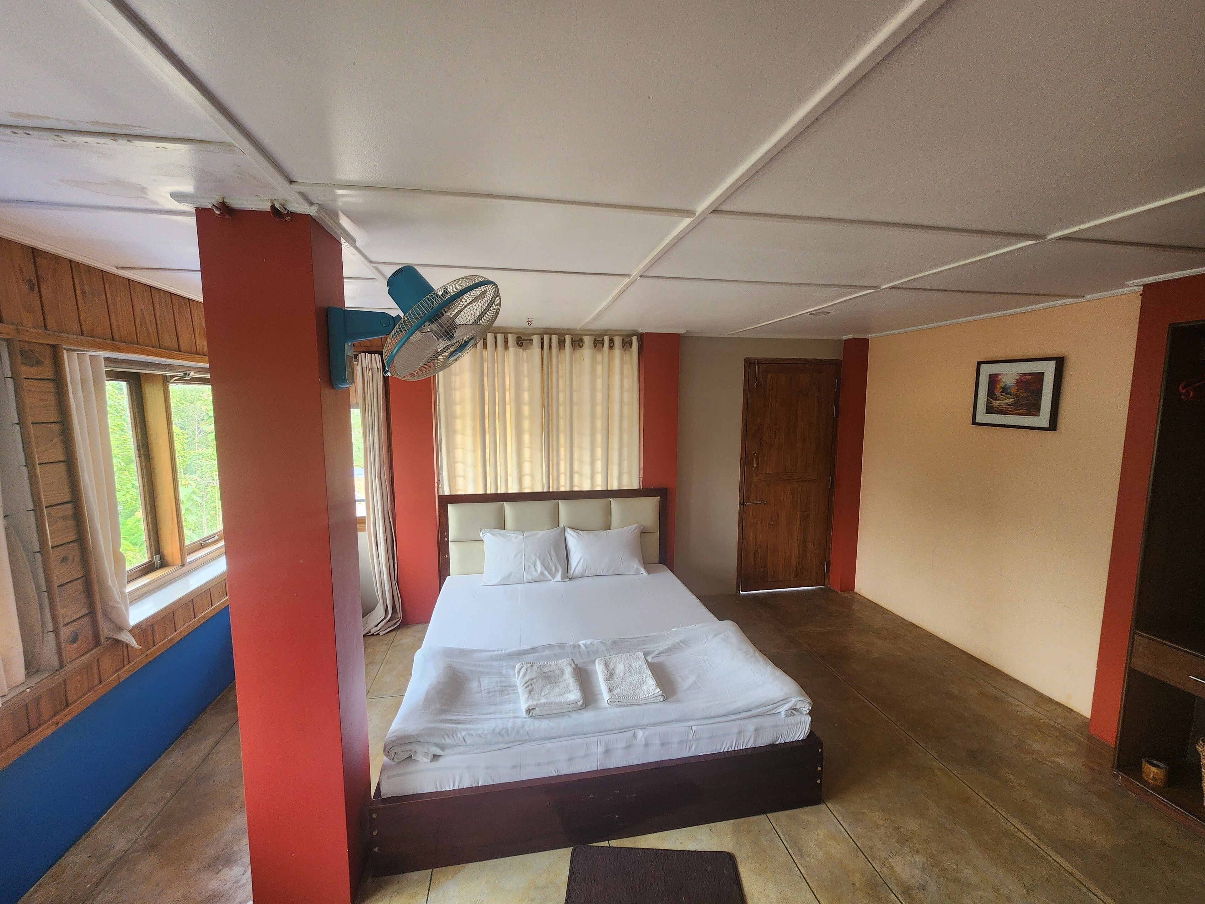 Uma cama de solteiro com roupa de cama branca, um ventilador de teto, uma janela com cortinas e uma porta de madeira.
