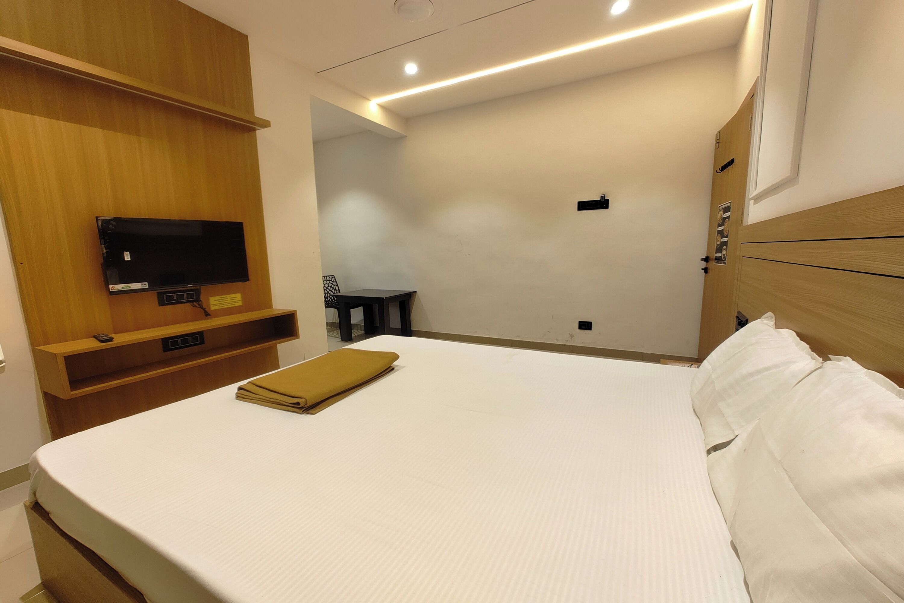 Quarto de hotel com uma cama, uma televisão em uma prateleira de madeira, uma mesinha e uma porta com moldura de madeira.