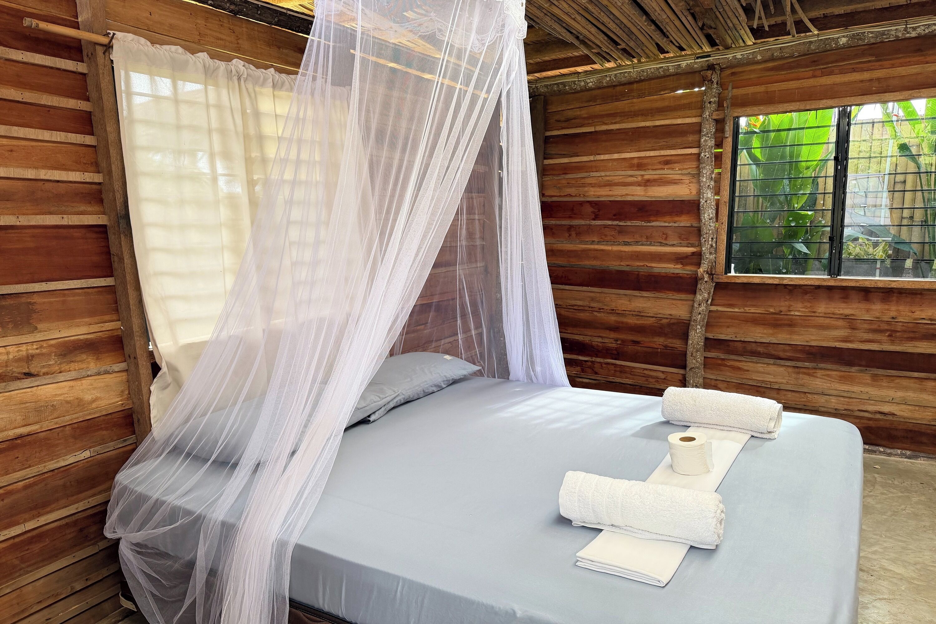 Quarto de cabana de madeira com cama, mosquiteiro e janela com vista para o verde.