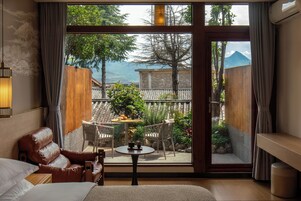 Quarto de hotel com uma grande janela, uma cama, área de estar com mesa e cadeiras, e vista para uma paisagem montanhosa.