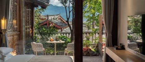 Quarto de hotel com vista para arquitetura chinesa tradicional através de uma porta de vidro, uma cama, uma mesa pequena com frutas e uma área de estar com cadeiras.