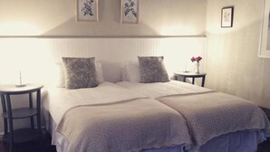 Uma cama de casal com colcha branca e duas almofadas, uma mesa de cabeceira com um vaso de flores e duas obras de arte florais emolduradas na parede.
