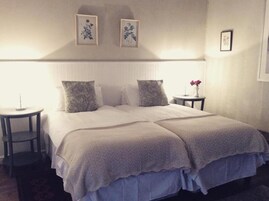 Uma cama de casal com colcha branca e duas almofadas, uma mesa de cabeceira com um vaso de flores e duas obras de arte florais emolduradas na parede.