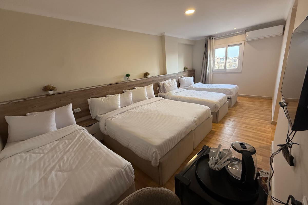Quarto de hotel com três camas, uma mesa com computador e janela com vista para a cidade.