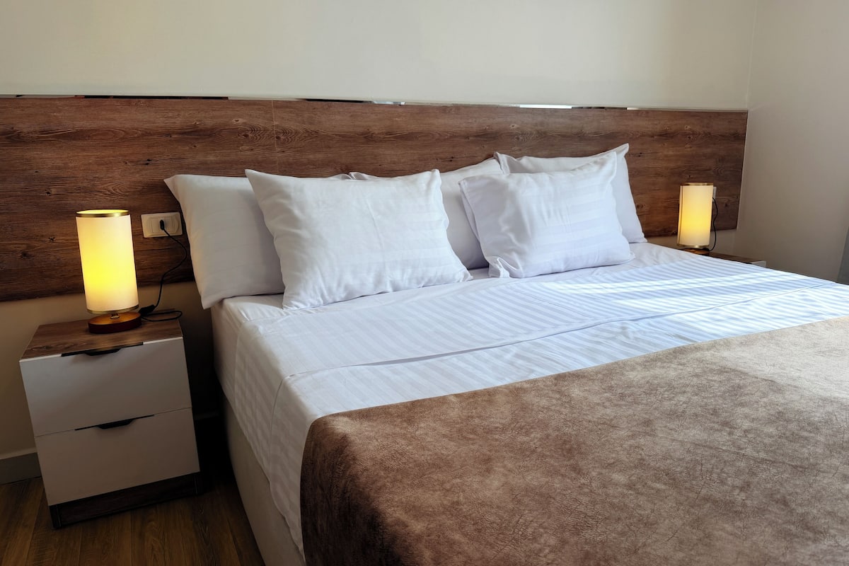 Uma cama bem arrumada com lençóis brancos e um cobertor marrom, com duas mesas de cabeceira com luminárias, em um quarto com cabeceira de madeira e piso de madeira.