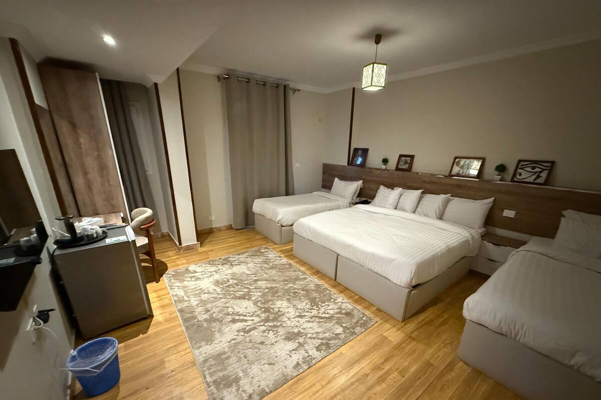 Quarto de hotel com uma cama grande, uma cama menor, uma escrivaninha com cadeira, um cesto de lixo azul e um tapete estampado.
