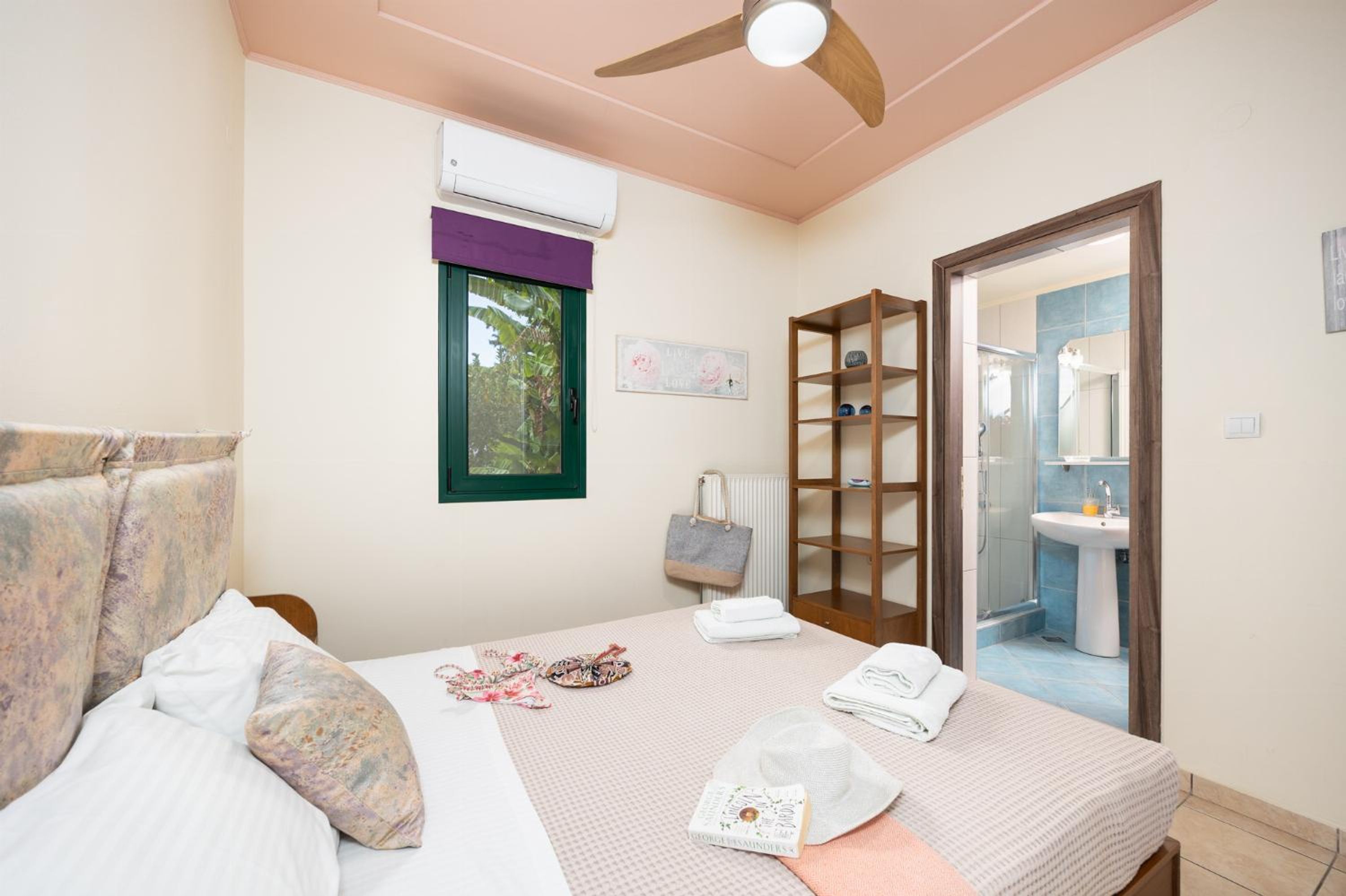 Quarto com cama, ventoinha no teto, janela com vista, guarda-roupa e banheiro visível por uma porta aberta.