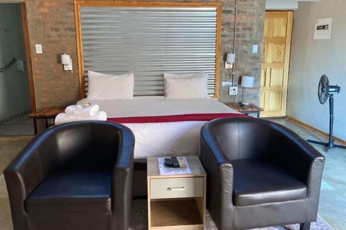 Quarto de hotel com uma cama grande, duas poltronas de couro, uma mesa de cabeceira com controle remoto e um ventilador.