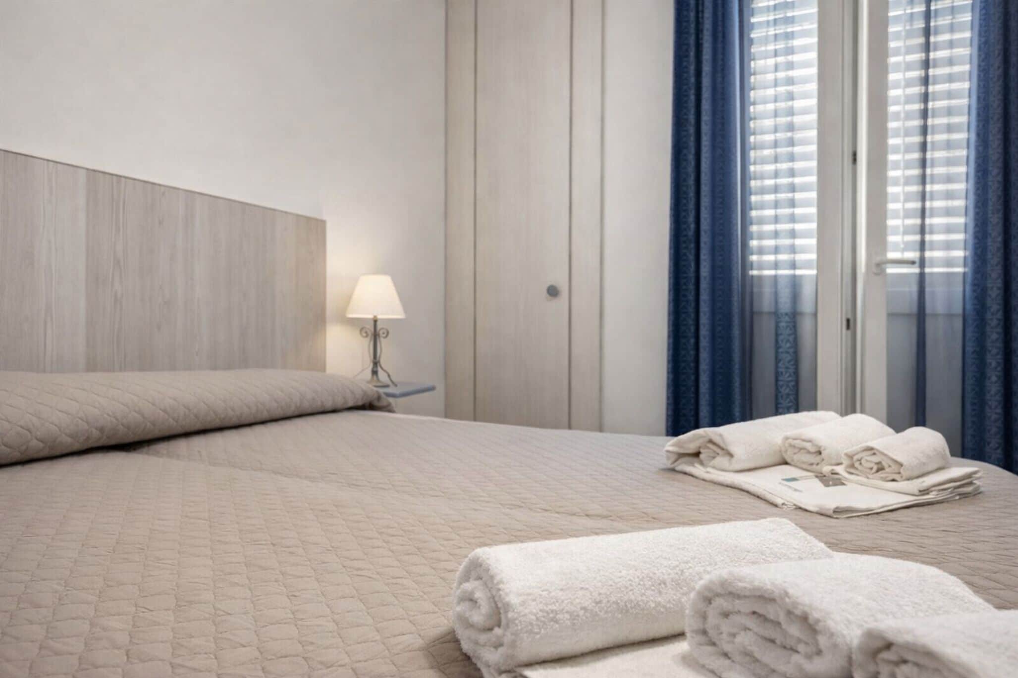 Quarto de hotel com uma cama, uma mesa de cabeceira com abajur, uma janela com cortinas e um conjunto de toalhas sobre a cama.