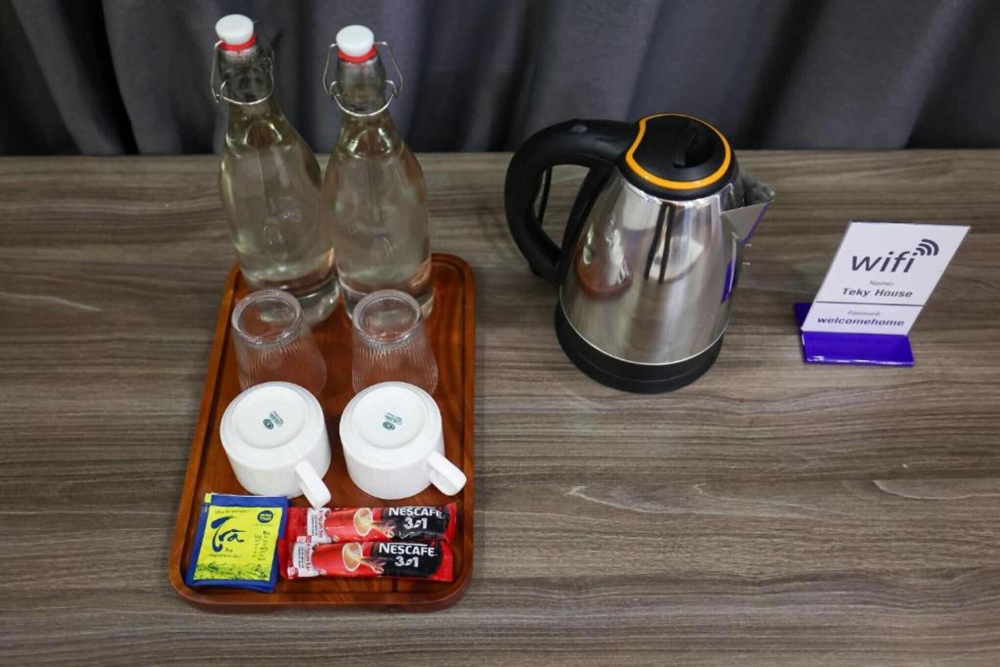Uma bandeja com bule, duas xícaras de chá, duas garrafas de água e dois sachês de Nescafé sobre uma mesa de madeira.