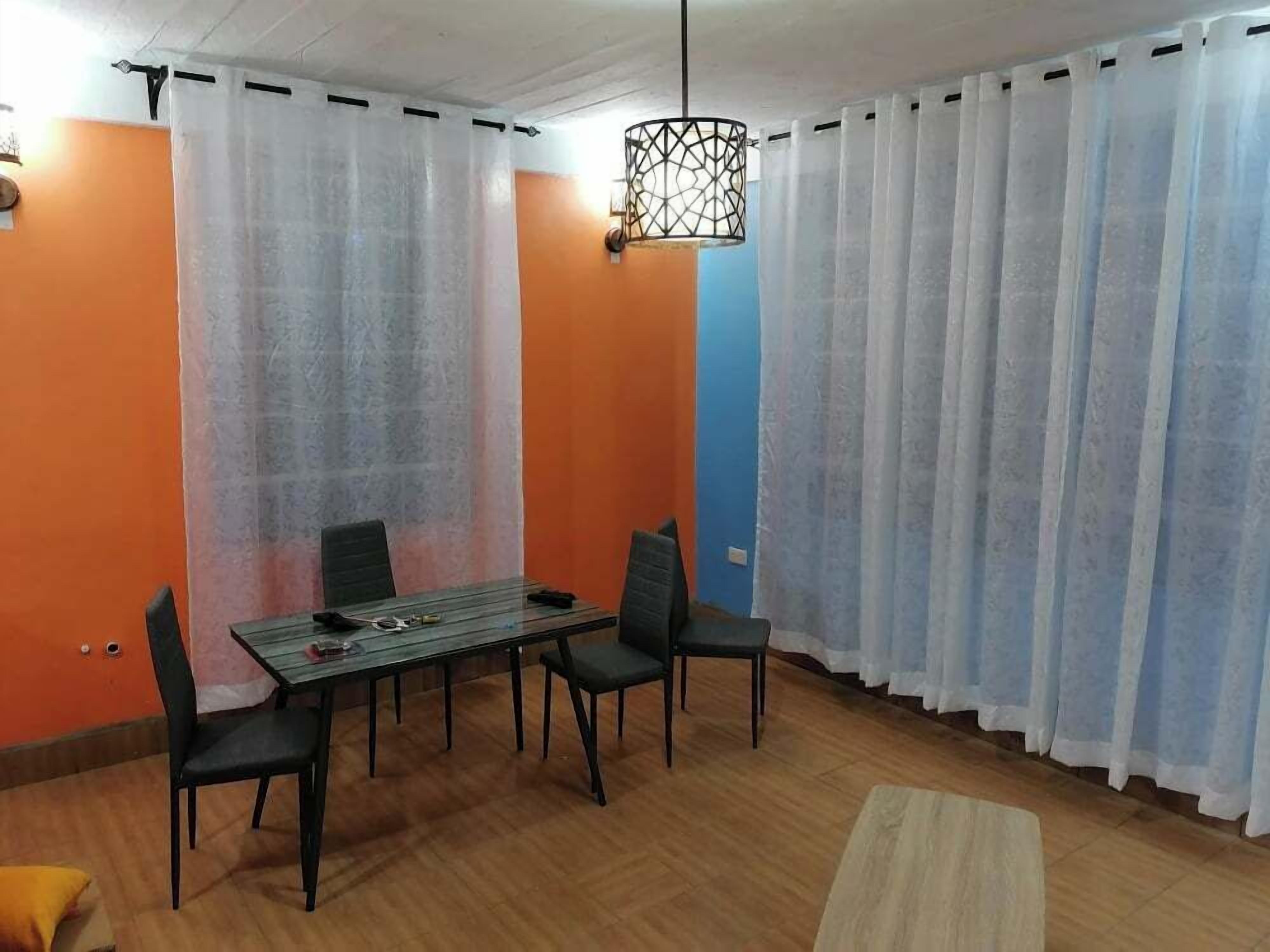 Quarto com mesa de madeira e quatro cadeiras, paredes laranja e azuis, e luminária pendente.