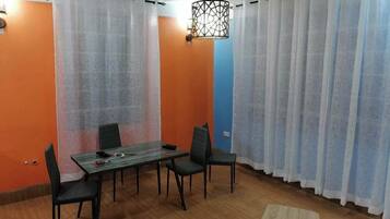 Quarto com mesa de madeira e quatro cadeiras, paredes laranja e azuis, e luminária pendente.