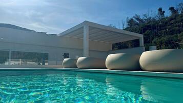 Piscina com água cristalina, um edifício moderno com fachada branca e uma área externa coberta com assentos.