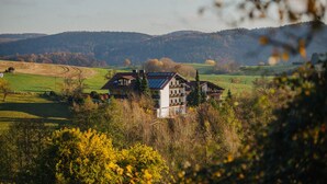 Uma casa grande com telhado de painéis solares, rodeada por uma paisagem verdejante e colinas ao fundo.