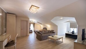 Ampla sala de estar com sofá, mesa de centro e televisão. Há também um espaço de jantar com mesa e cadeiras. O cômodo possui piso de madeira e um lustre.
