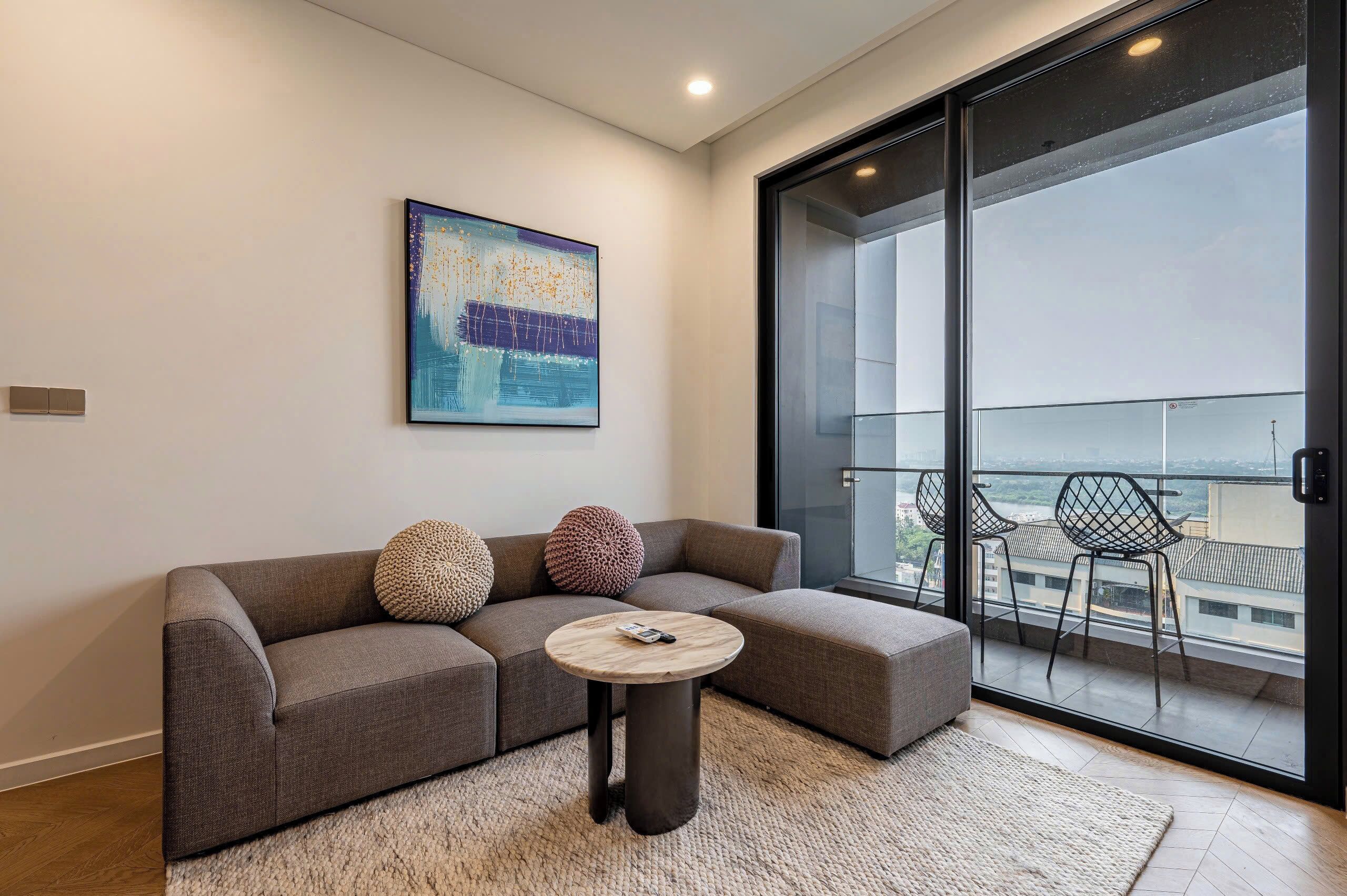Uma sala de estar moderna com um sofá modular cinza, uma mesa de centro redonda de madeira e uma grande janela com vista para a cidade.