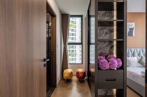 Quarto de hotel moderno com porta de madeira, vista de um edifício pela janela, prateleira de mármore com flores roxas e cama com cabeceira cinza.