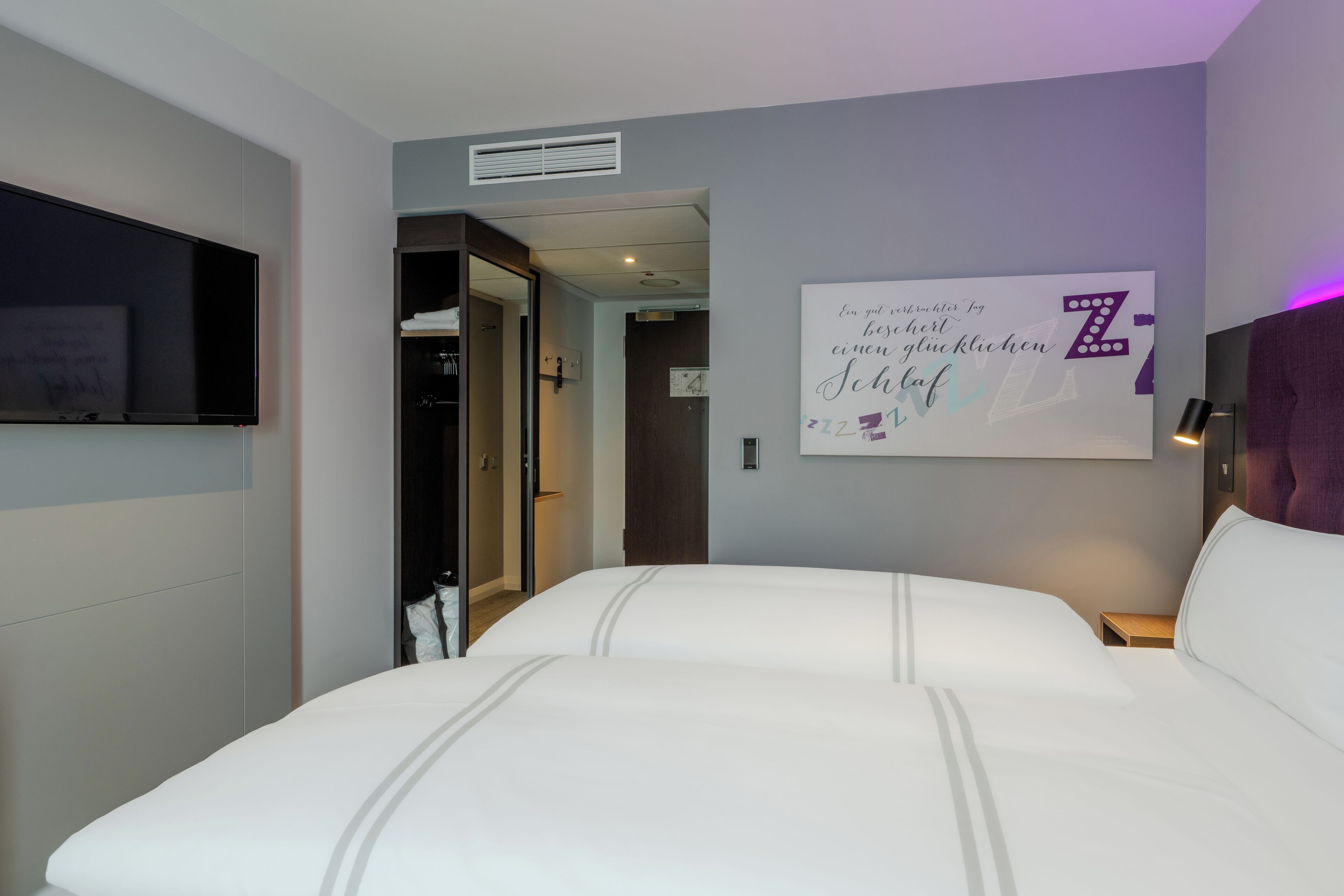 Quarto de hotel com uma cama grande, televisão de tela plana, obra de arte na parede e um corredor que dá para um banheiro.