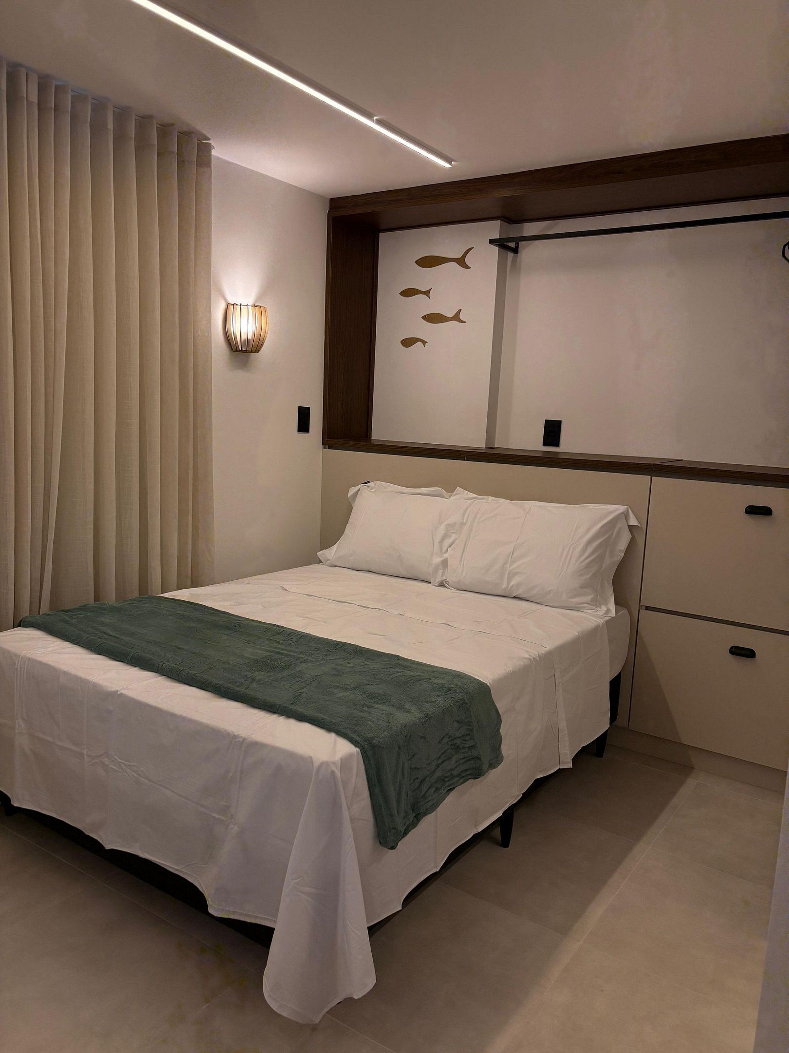 Quarto de hotel com cama, mesa de cabeceira, luminária na parede e janela com cortinas.