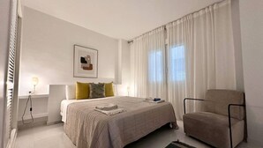 Quarto com cama, poltrona, mesa de cabeceira e janela com cortinas.