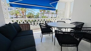 Um pátio externo com uma mesa redonda e cadeiras pretas, um toldo azul e um corrimão branco.