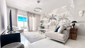 Uma sala de estar moderna com um sofá branco, uma mesa de centro, uma TV em um móvel e uma grande janela com cortinas.