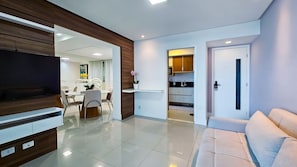Uma sala de estar moderna com televisão de tela plana, sofá, área de refeições com mesa e cadeiras, e cozinha visível por uma porta aberta.
