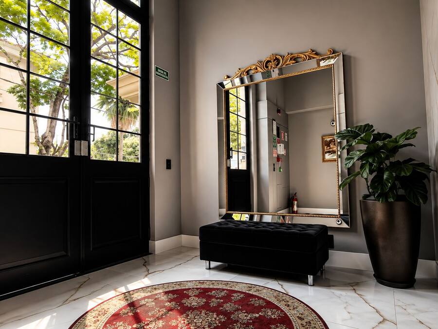 Um corredor com uma porta preta, um espelho grande, um banquinho preto, um tapete vermelho estampado e uma planta em vaso.