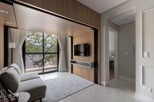 Uma sala de estar moderna com sofá, televisão de tela plana fixada em uma parede de madeira, uma janela grande com cortinas, e um corredor que leva a outro cômodo.