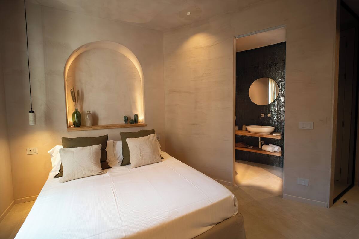 Quarto moderno com uma cama grande, um arco embutido sobre o espelho e um banheiro com espelho redondo e pia.