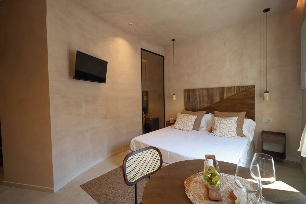 Quarto de hotel moderno com uma cama grande, uma televisão de tela plana, uma mesa de jantar com taças de vinho e uma cadeira.