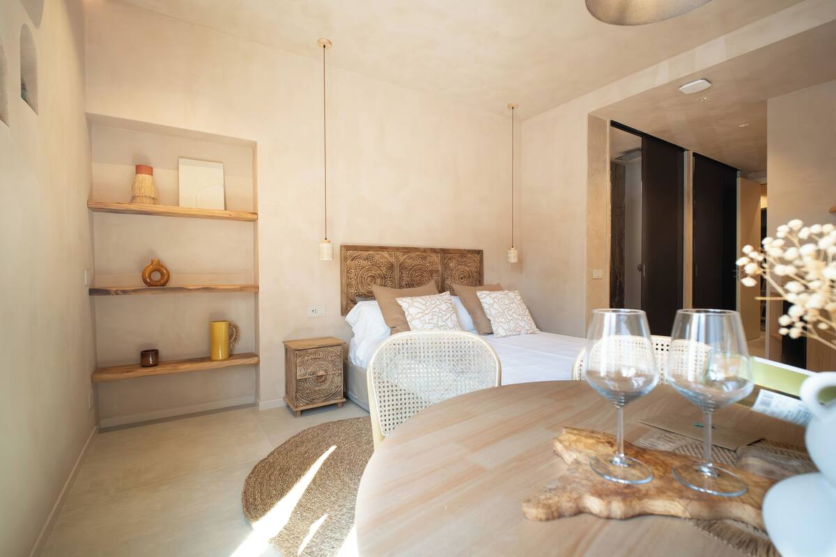 Quarto moderno com cabeceira de madeira, mesa de cabeceira com um vaso e duas taças de vinho, e uma janela grande.