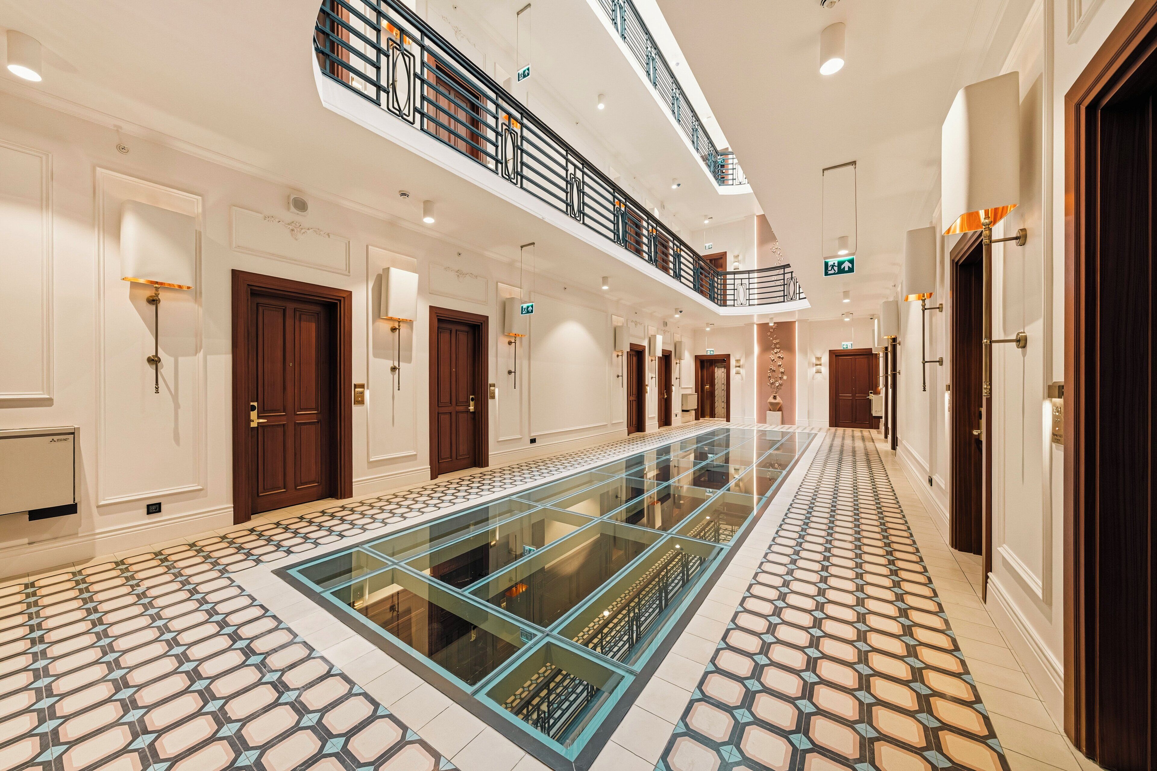 Um hall de hotel espaçoso com piso de vidro, várias portas e uma varanda no segundo andar.