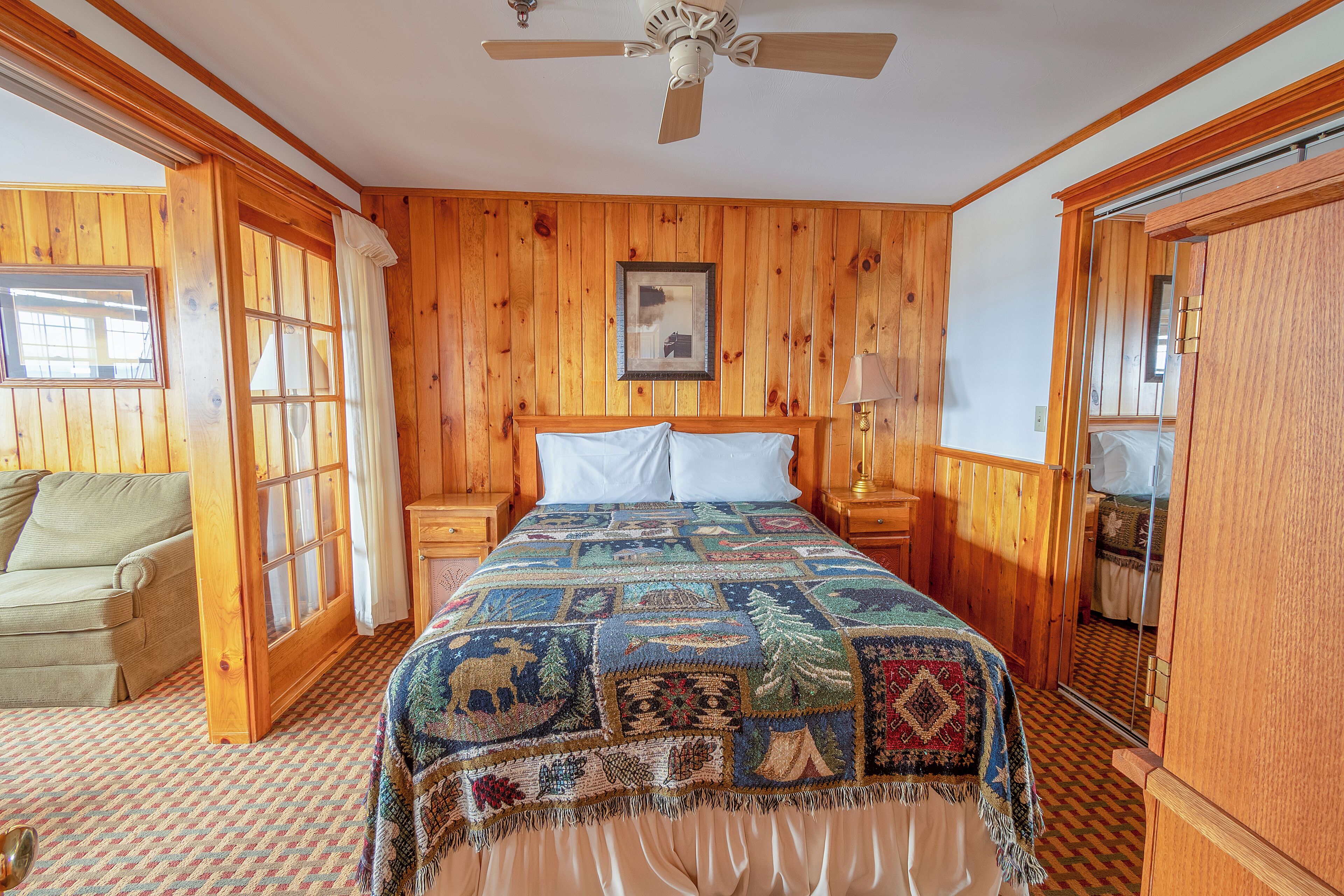 Quarto com painéis de madeira, uma cama, uma mesa de cabeceira, uma luminária e uma janela com cortinas.