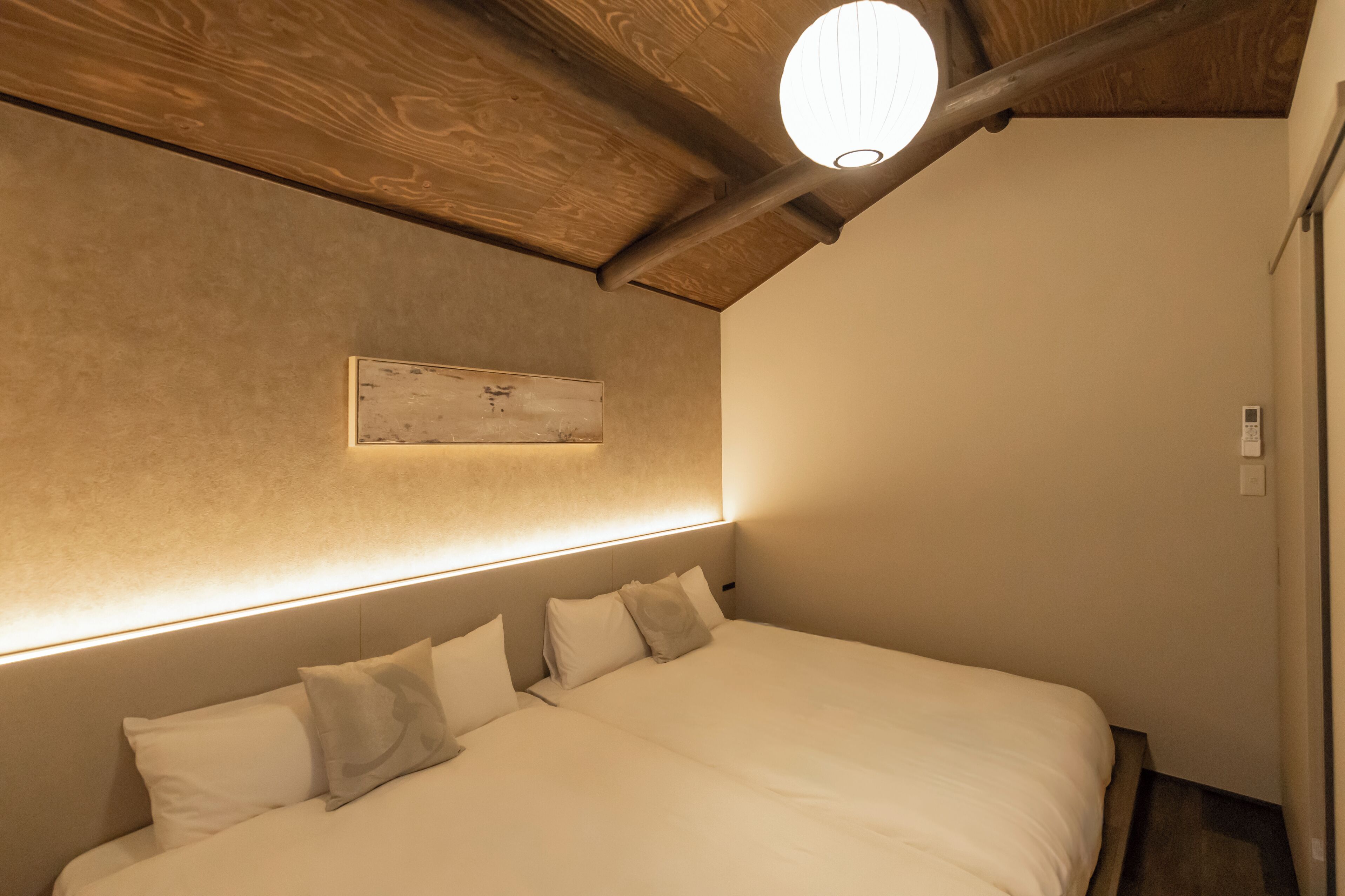 Um quarto com uma cama grande, teto de madeira, luminária fixada na parede e uma pequena pintura.