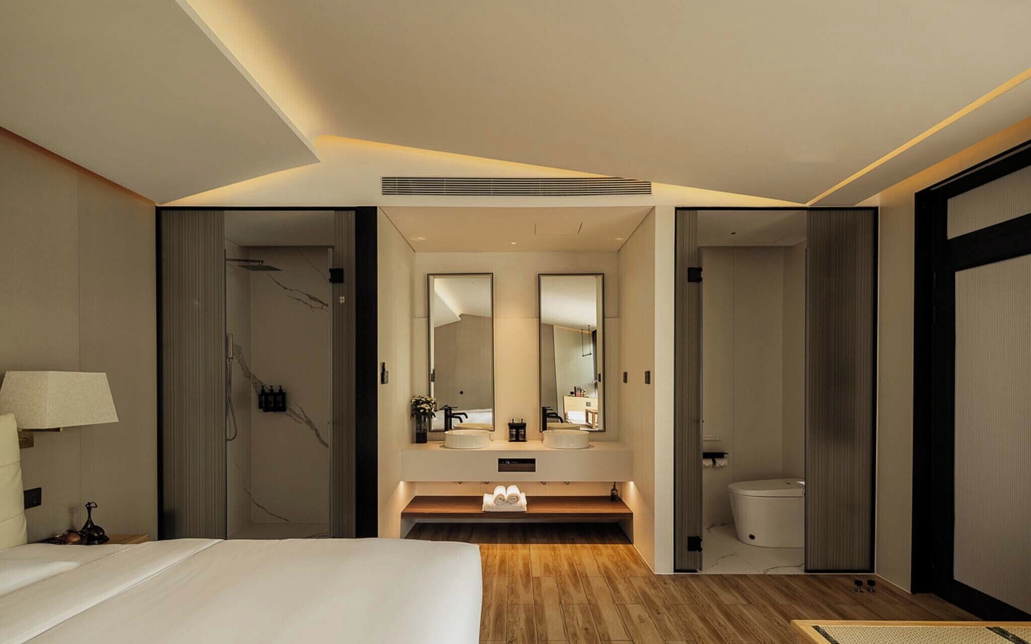 Quarto de hotel moderno com uma cama grande, banheiro com vaso sanitário e chuveiro, e toucador com espelho.