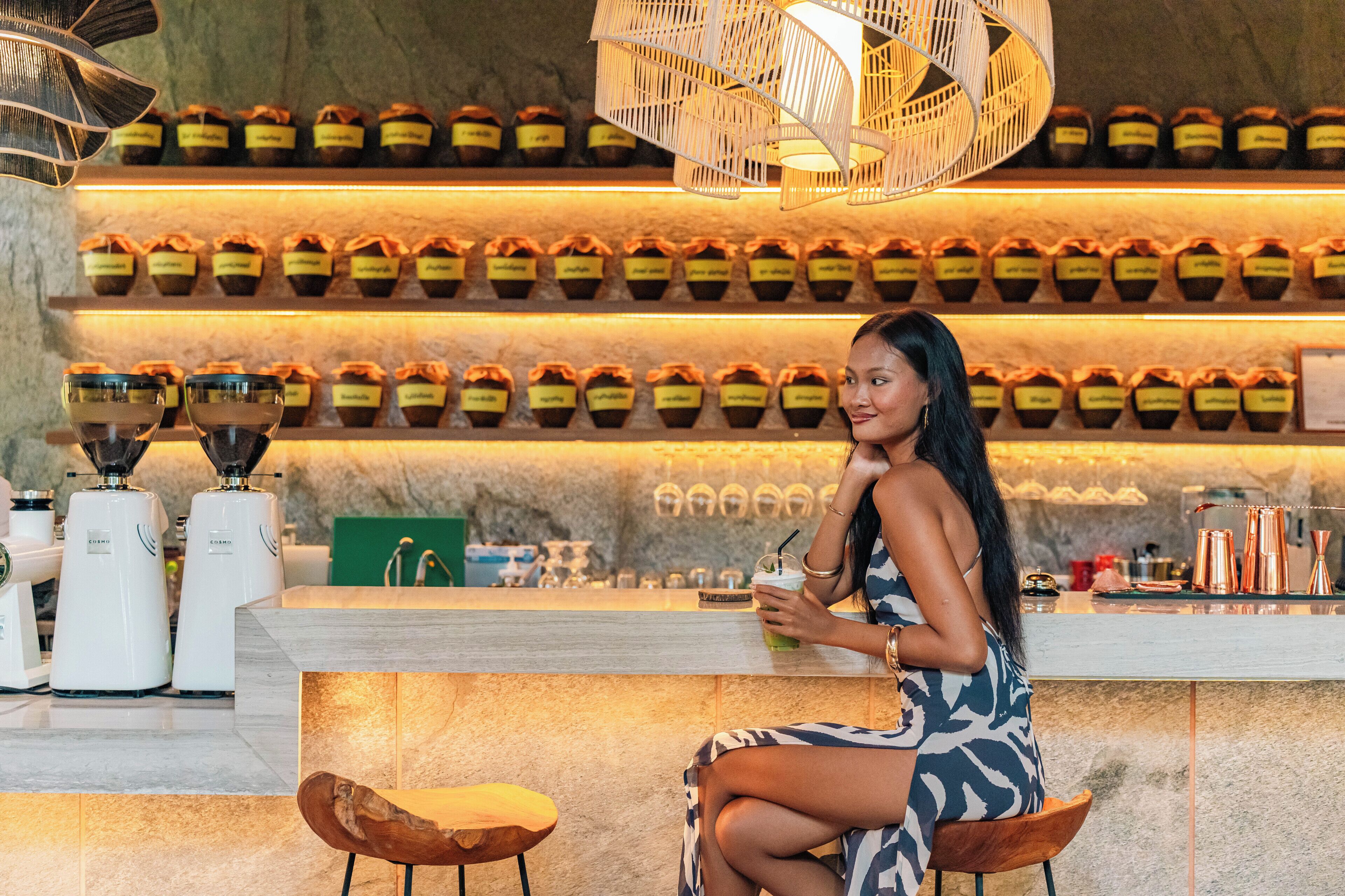 Uma área de bar com uma mulher sentada, um balcão com diversos objetos e prateleiras com potes.