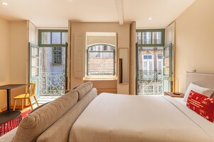 Quarto de hotel com cama, sofá, mesinha e cadeira. Janela com vista para edifícios.