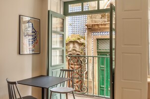 Quarto com mesa e cadeiras, quadro pendurado na parede e vista de um edifício com fachada azul e branca através de uma porta aberta.