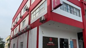 Um hotel de vários andares com fachada em vermelho e branco, um letreiro chamativo 'HOTEL MIDAH' e uma entrada ampla com porta de vidro.