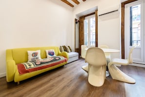 Um sofá amarelo vibrante com uma manta vermelha e preta, uma cadeira branca e uma mesa pequena em um quarto com piso de madeira e paredes brancas.