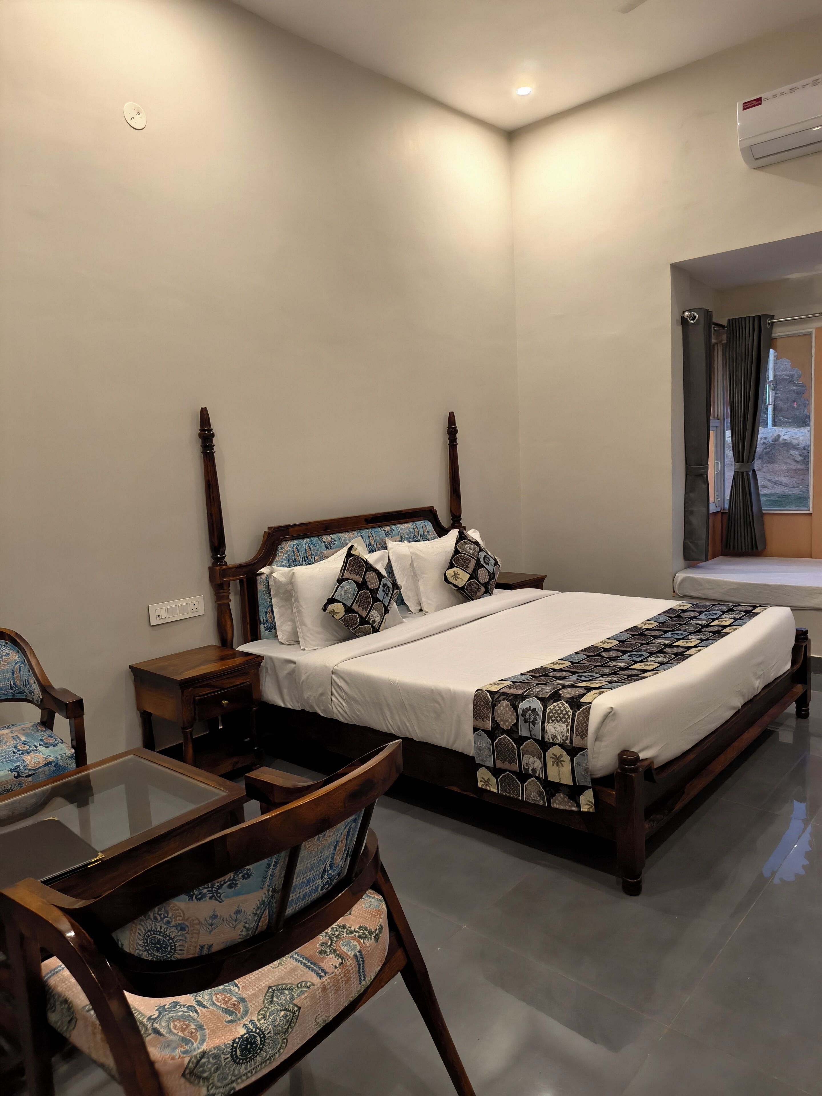 Quarto com uma cama grande, cabeceira de madeira e colcha estampada. Há uma mesinha de cabeceira, uma cadeira com almofada estampada e uma janela com cortinas.